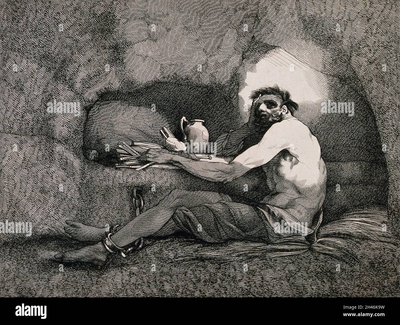 A prisoner is sitting on straw in a cave with his feet chained to the ...