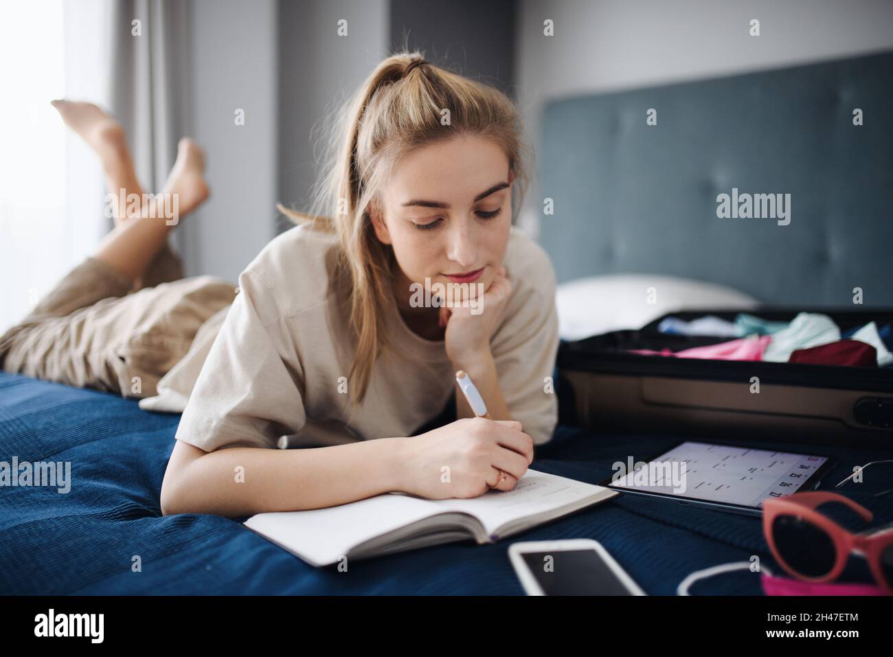 Portrait of young woman writing notes and packing clothes into suitcase ...