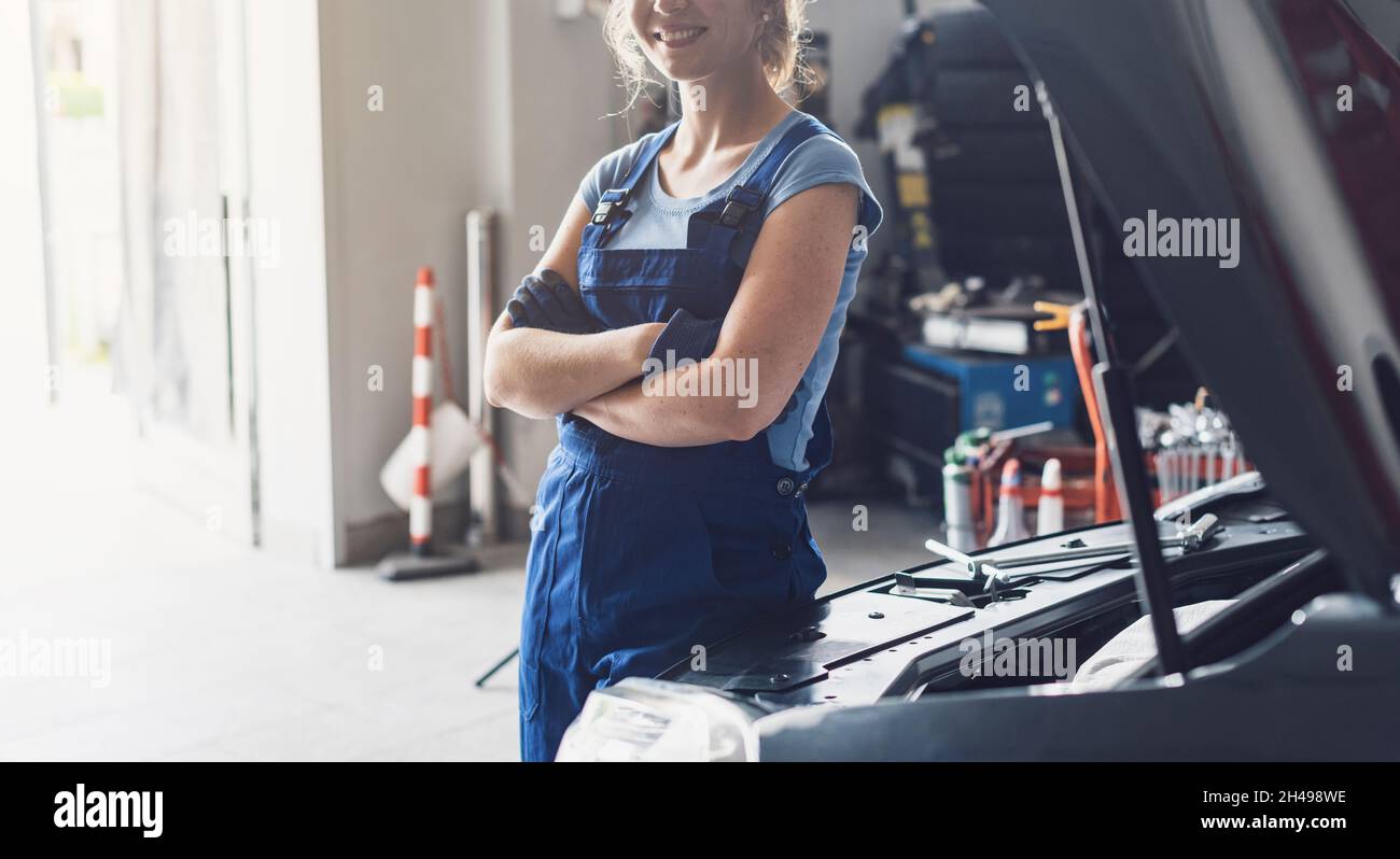 Smiling female mechanic posing crossed next to a car with hood up ...