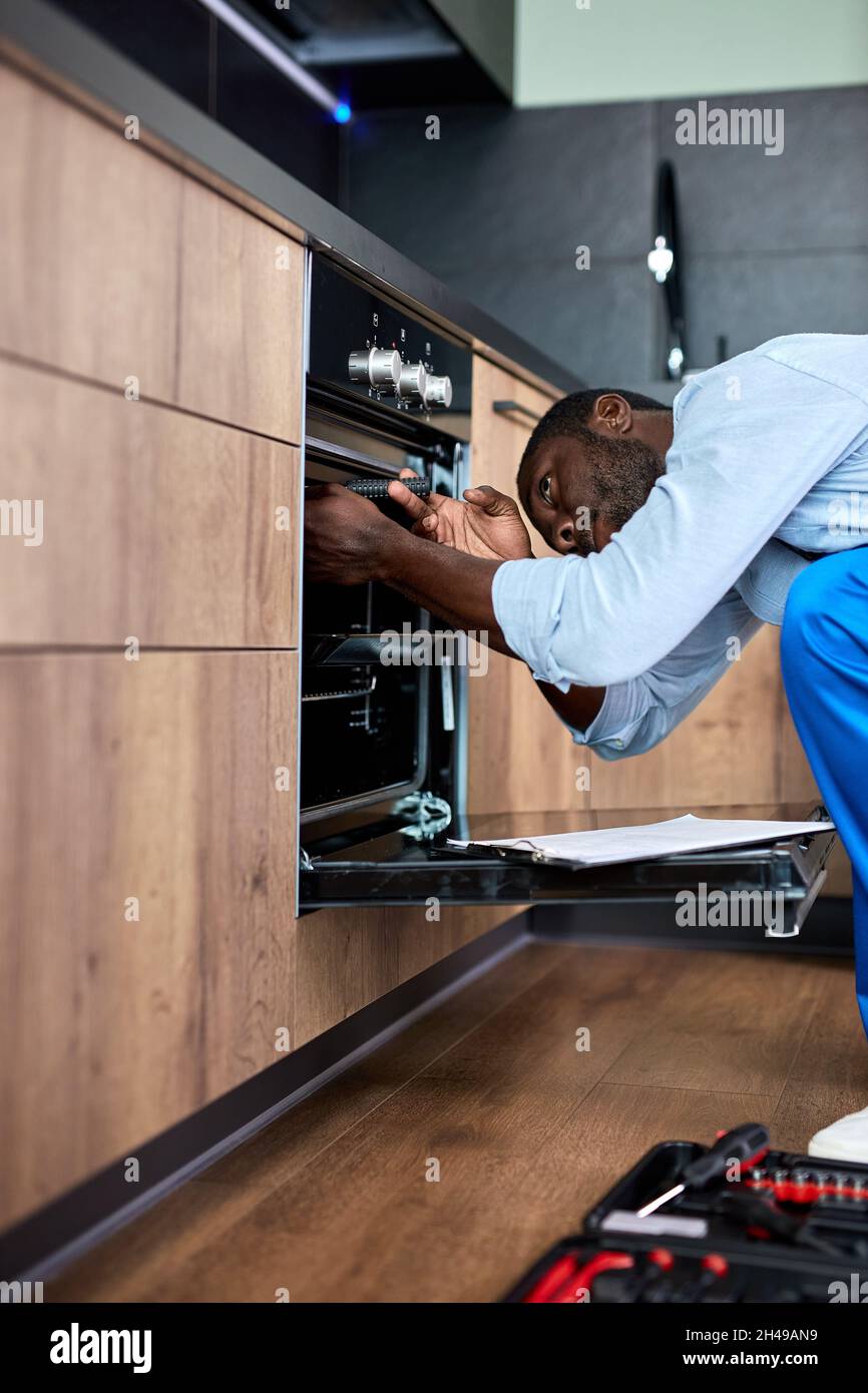 Afro american repairman in uniform working, fixing broken oven in ...