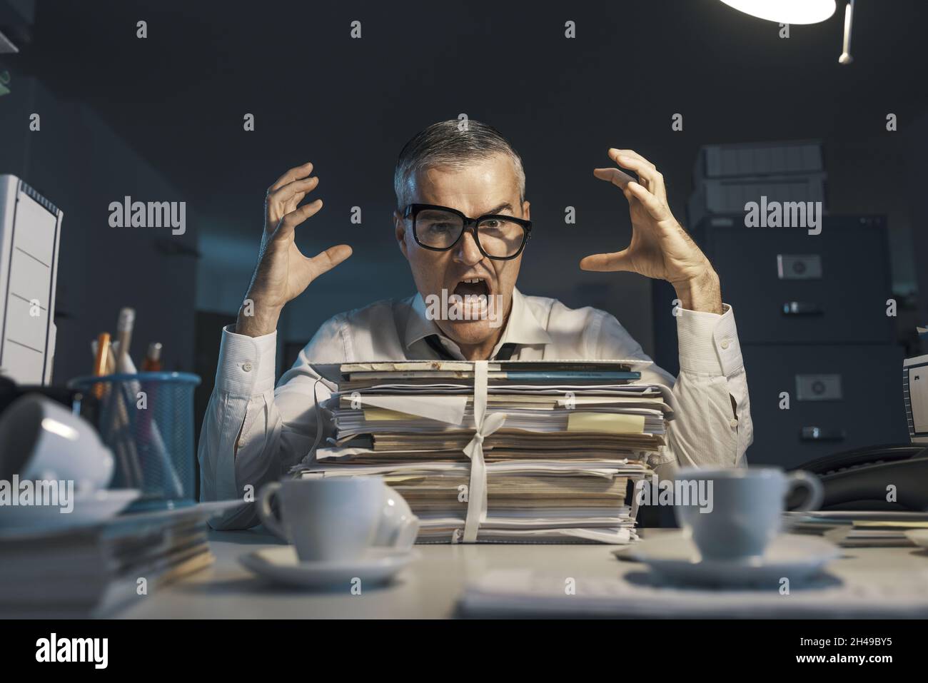 Angry businessman working overtime late at night surrounded by piles of ...
