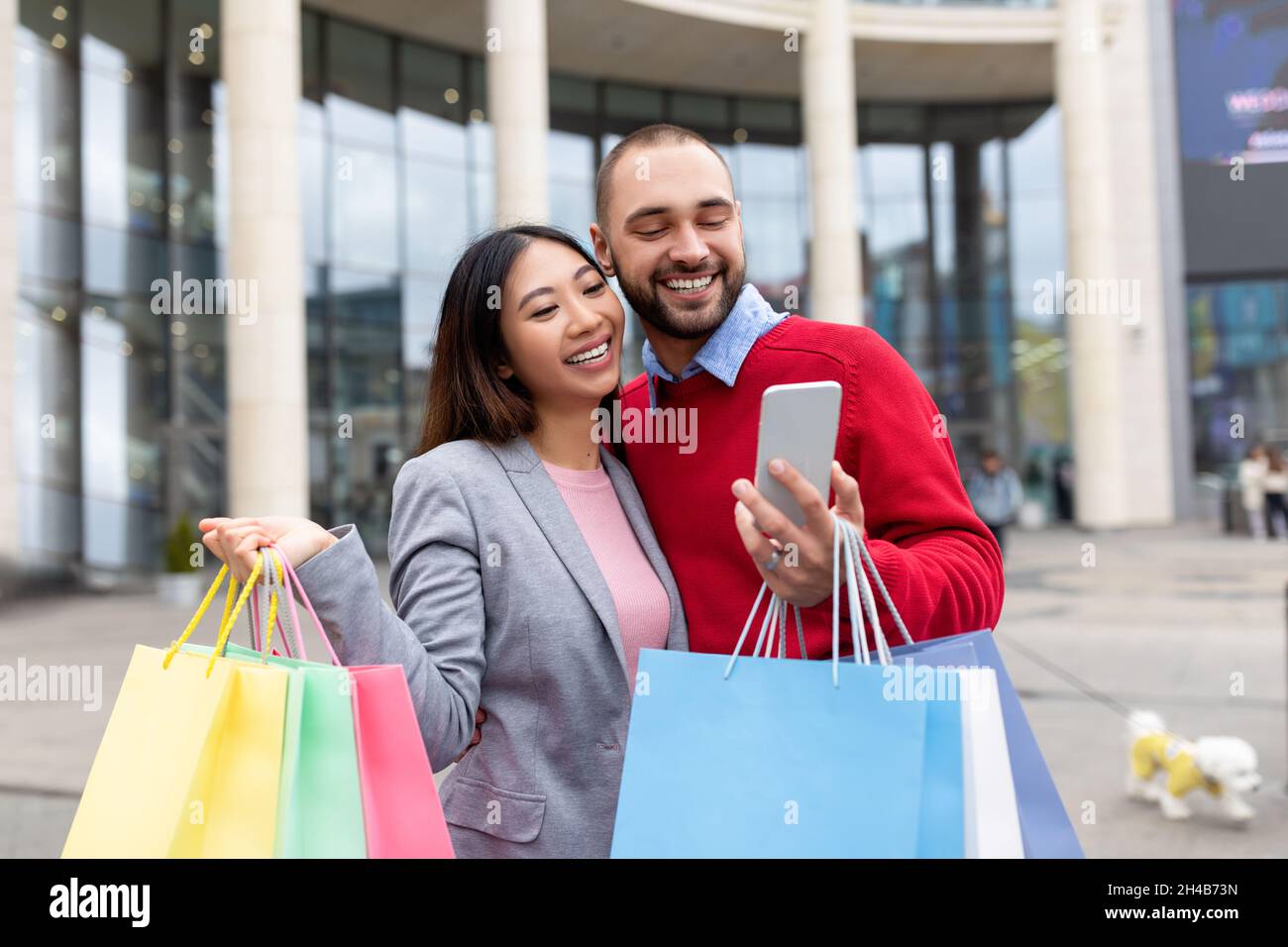 Positive multiracial couple with bright gift bags and cellphone placing ...