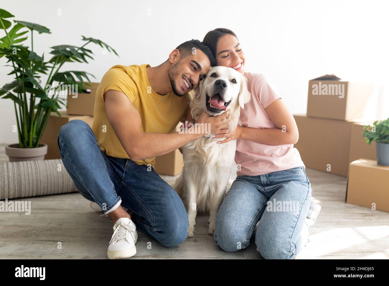 Happy young diverse couple hugging their cute dog, sitting on floor ...