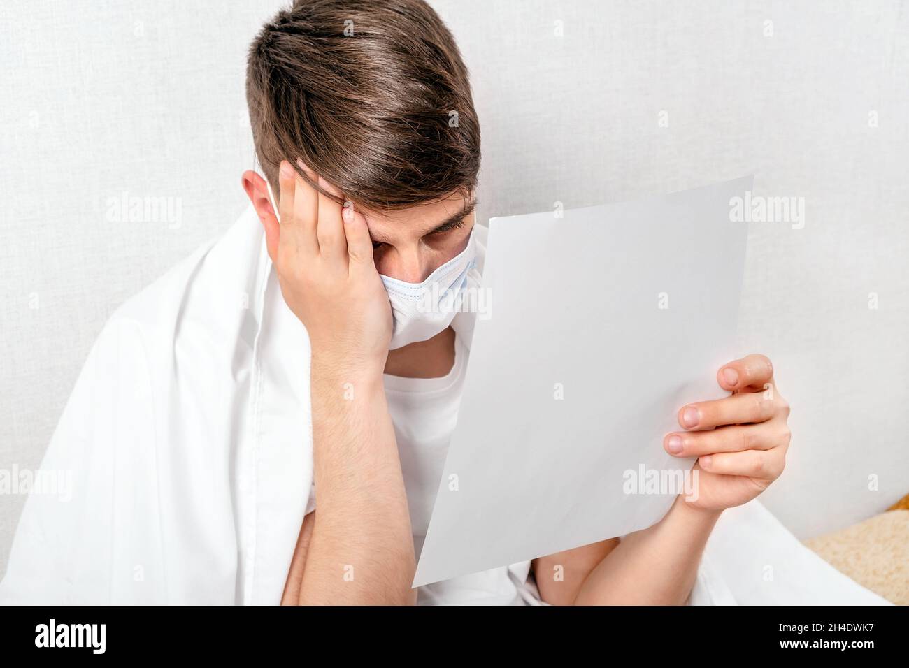 Troubled Young Man in Flu Mask read a Document by the White Wall at the ...