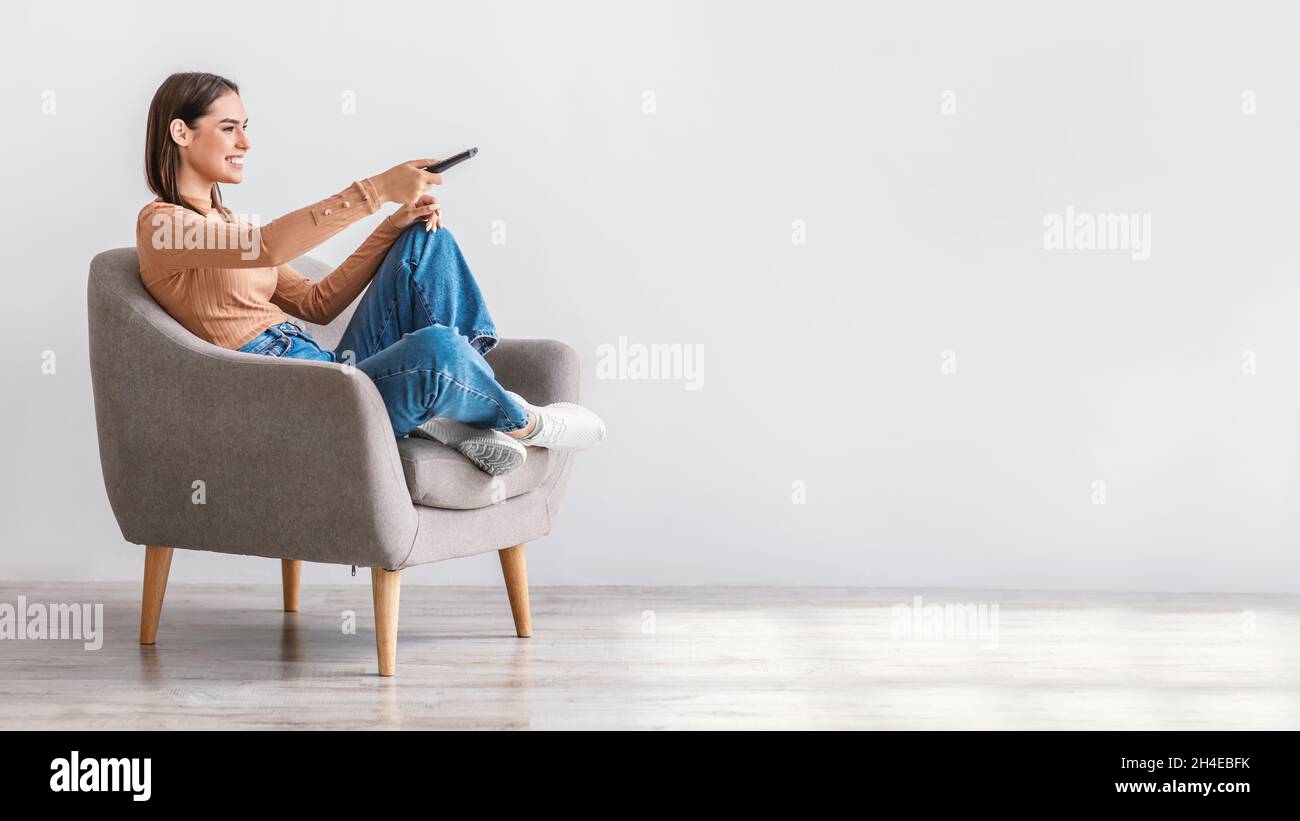 Young Caucasian lady with remote control sitting in armchair and ...