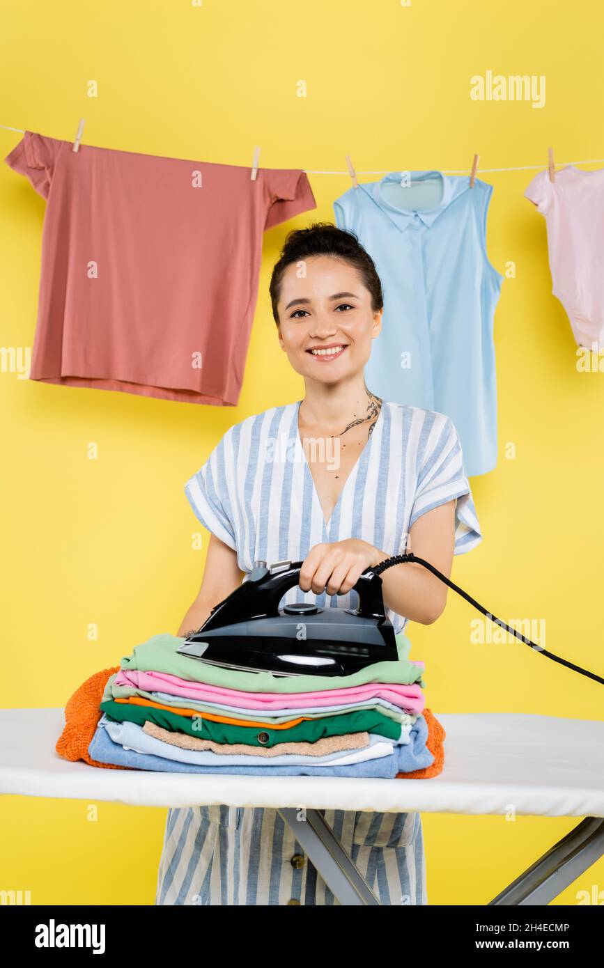 pleased woman holding iron near stack of clothes on ironing board on ...