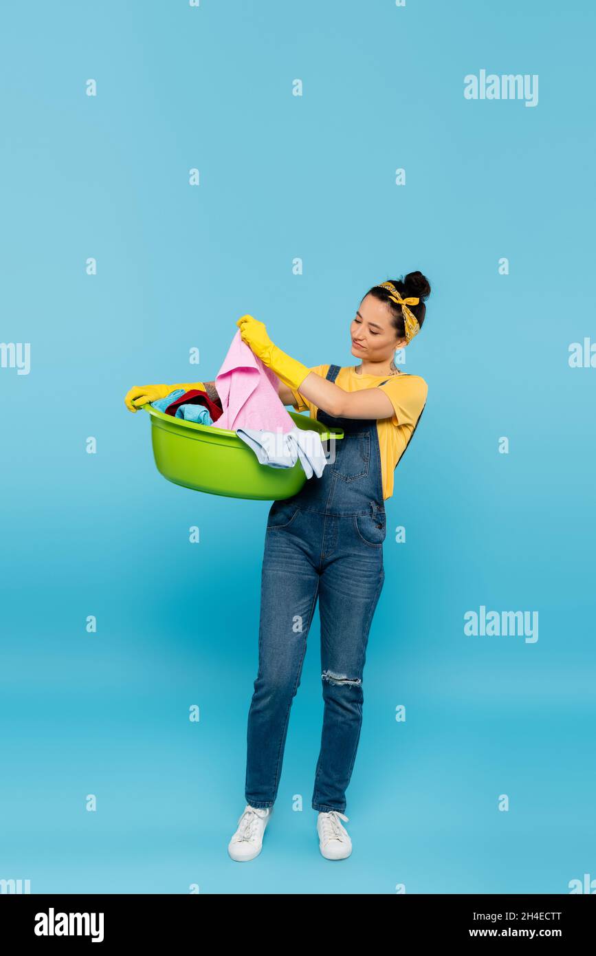 full length view of housewife in denim overalls holding laundry bowl ...