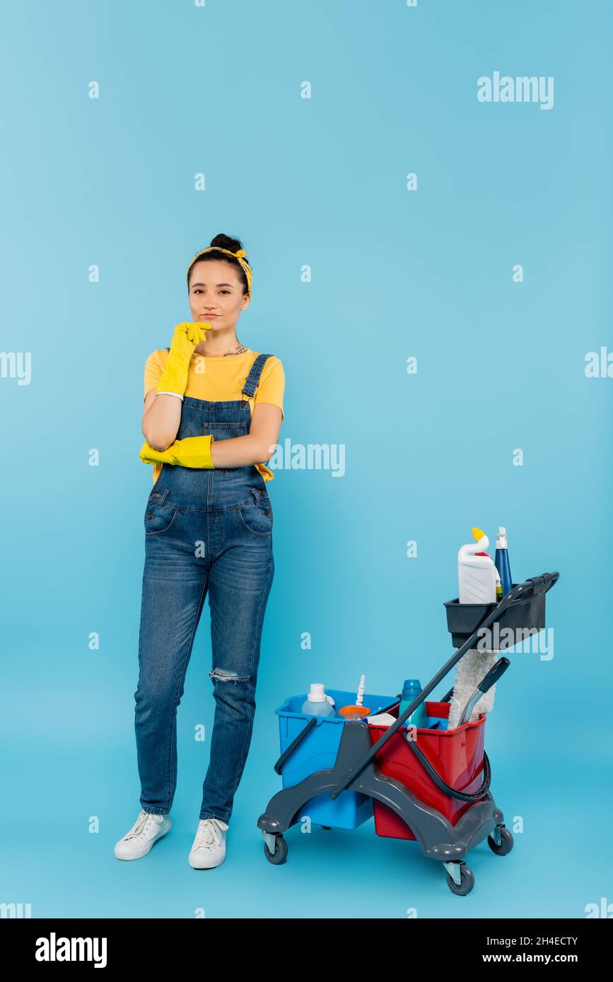 cleaner in rubber gloves and denim overalls looking at camera near cart ...