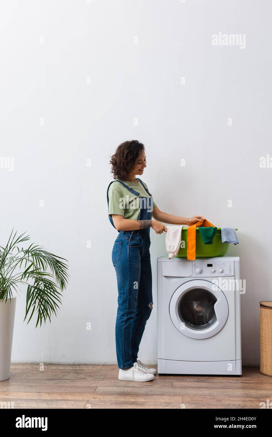 housewife in denim overalls standing near laundry bowl on washing ...