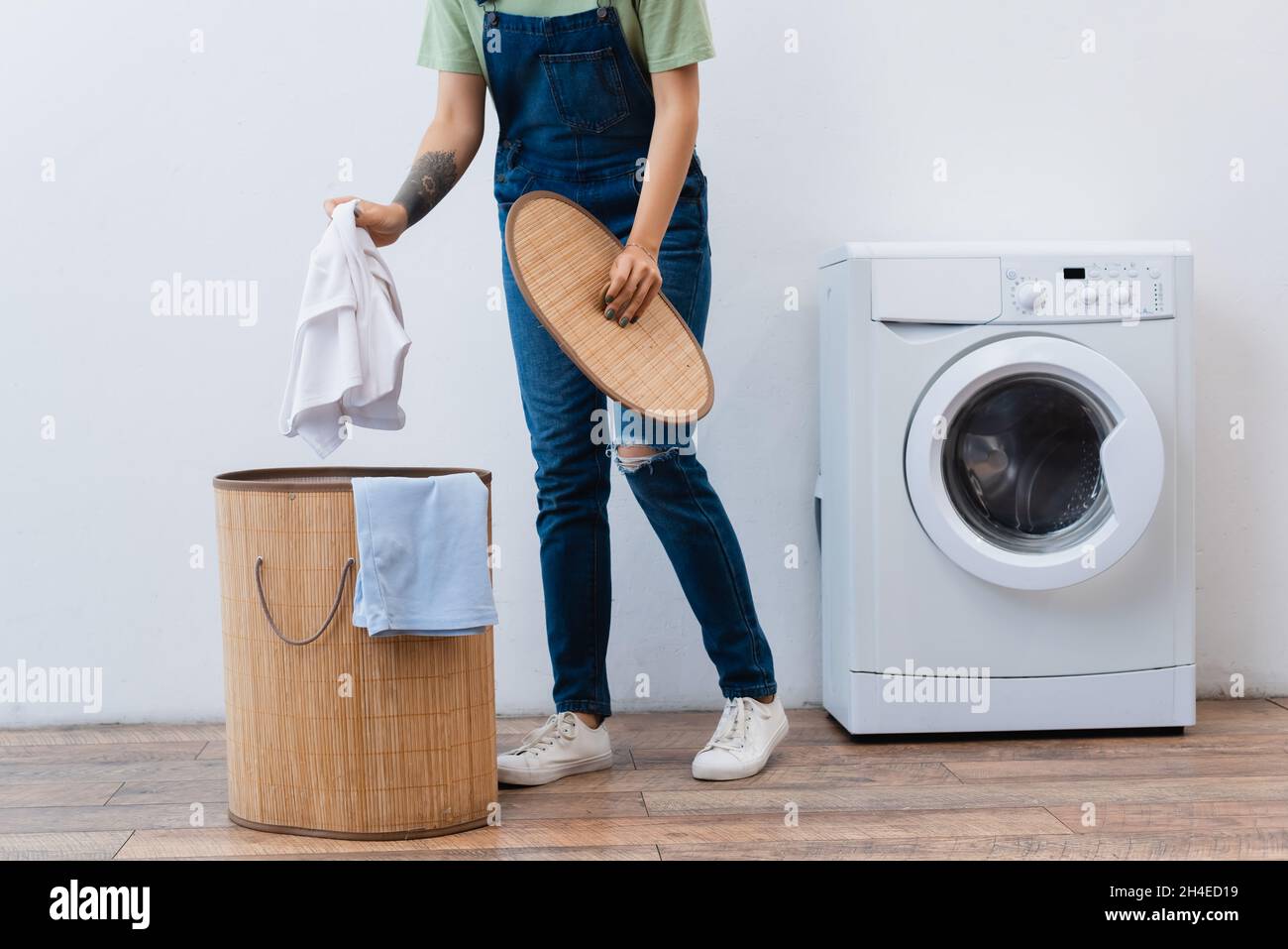 partial view of woman in denim overalls holding clothes near laundry ...