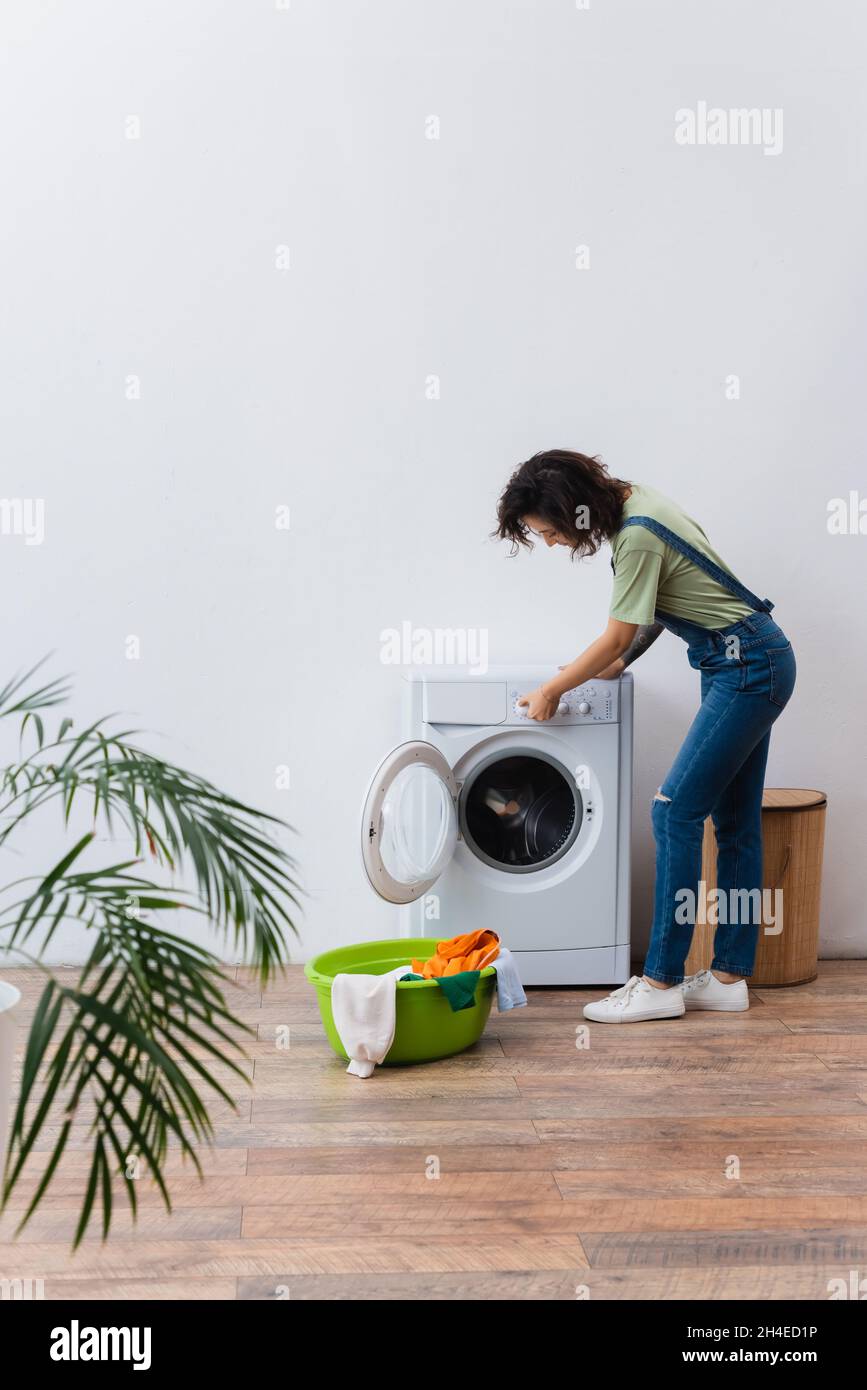 brunette woman operating washing machine near clothes in laundry bowl ...