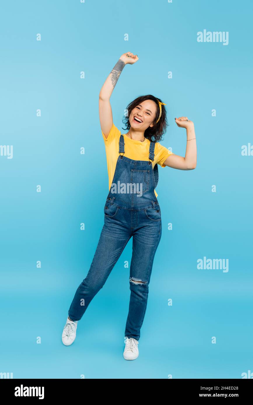 full length view of excited woman in denim overalls showing triumph ...