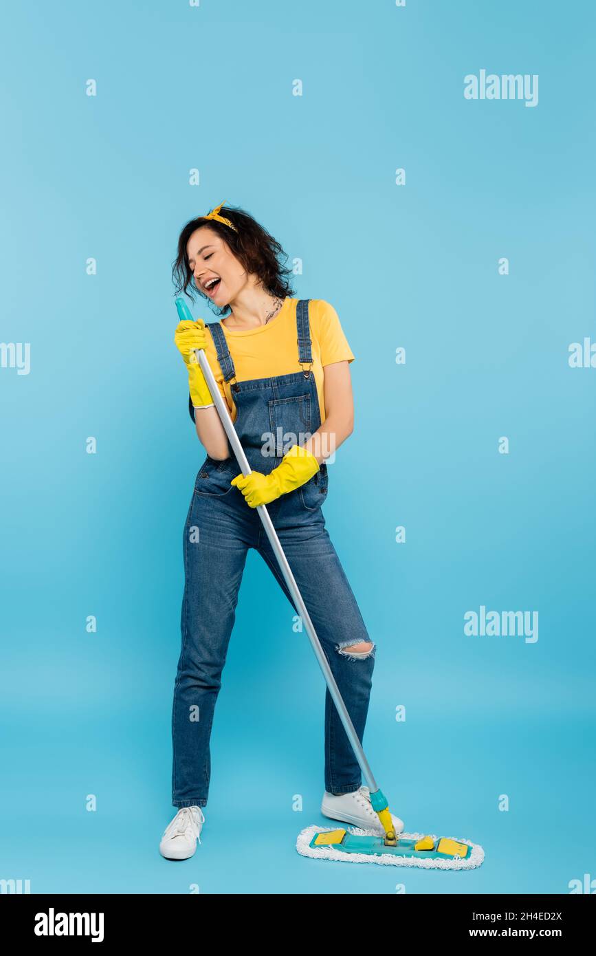 cheerful housewife in denim overalls and rubber gloves singing with mop ...