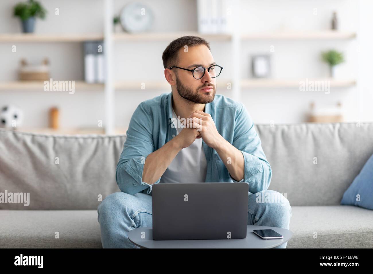 Serious young Caucasian guy sitting on couch in front of laptop ...
