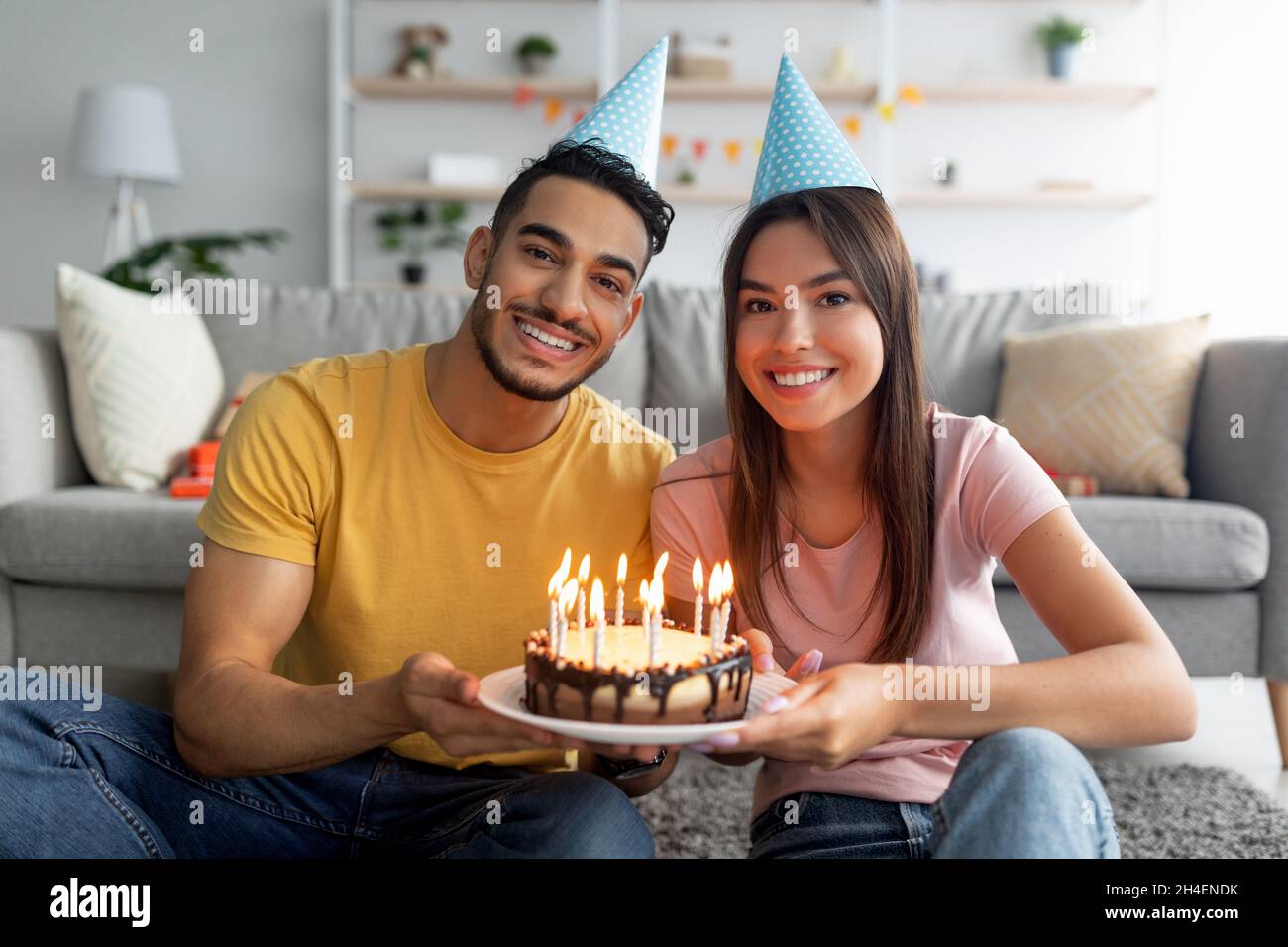 Cheery millennial multiracial couple in party hats holding birthday ...
