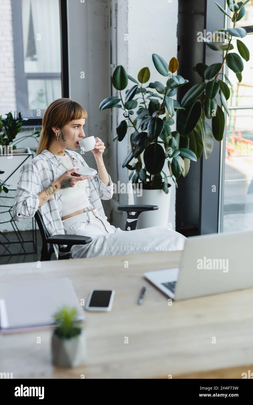 tattooed businesswoman drinking coffee while sitting near work desk and ...