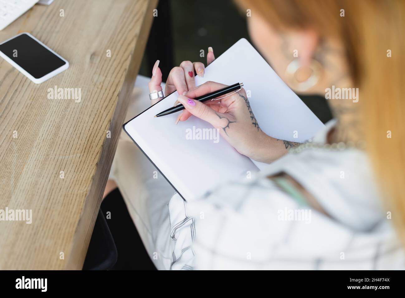 cropped view of blurred woman with tattoo writing in notebook near ...