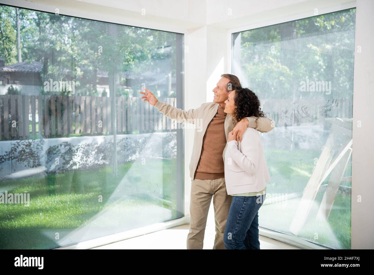 cheerful man pointing with hand at panoramic windows while hugging wife ...
