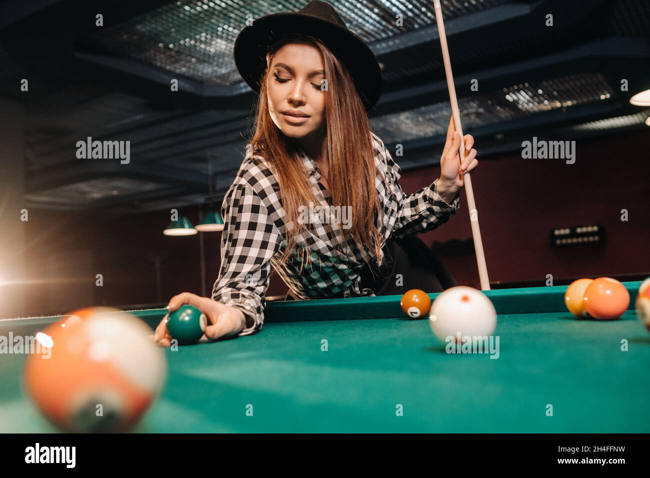 A girl in a hat in a billiard club with a cue and balls in her hands ...
