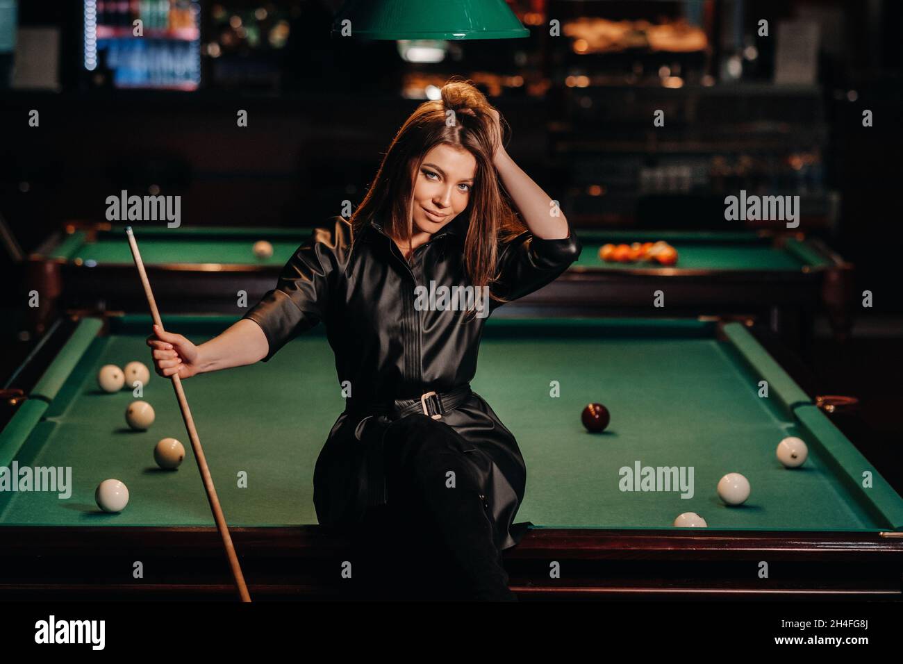 A girl with a cue in her hands sits on a table in a billiard club ...