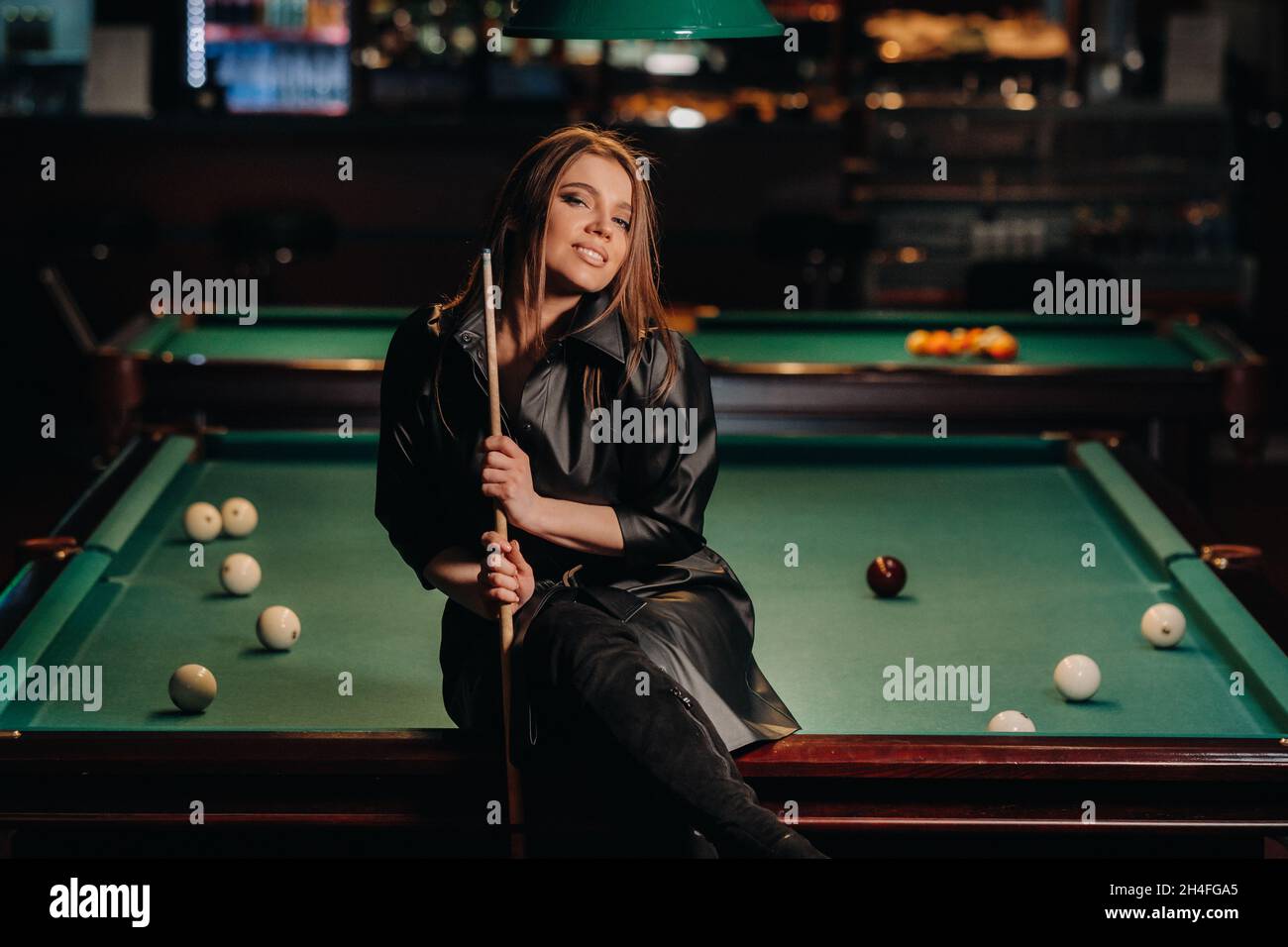 A girl with a cue in her hands sits on a table in a billiard club ...