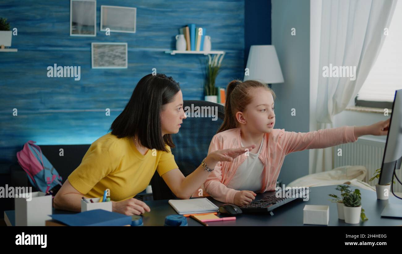 Parent and little girl doing homework together at desk and using computer with keyboard for ...