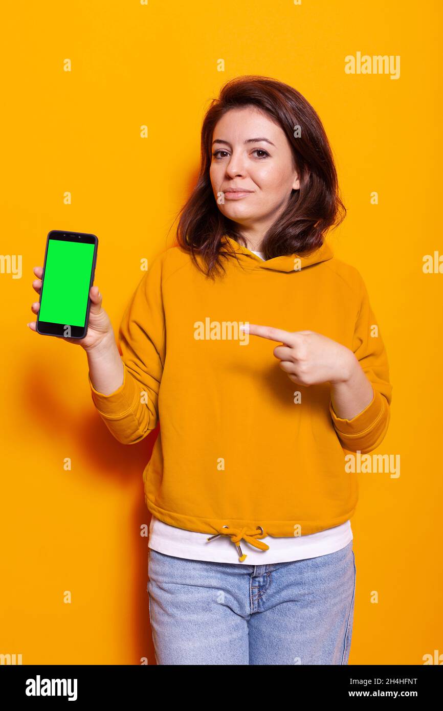 Woman pointing at smartphone with vertical green screen in studio ...