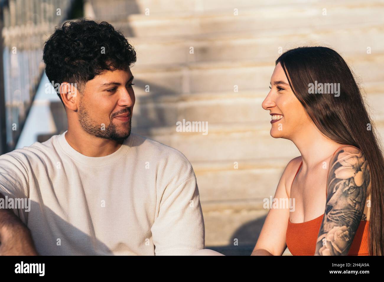 Two friends smiling while talking on an outdoor stairs during sunset ...