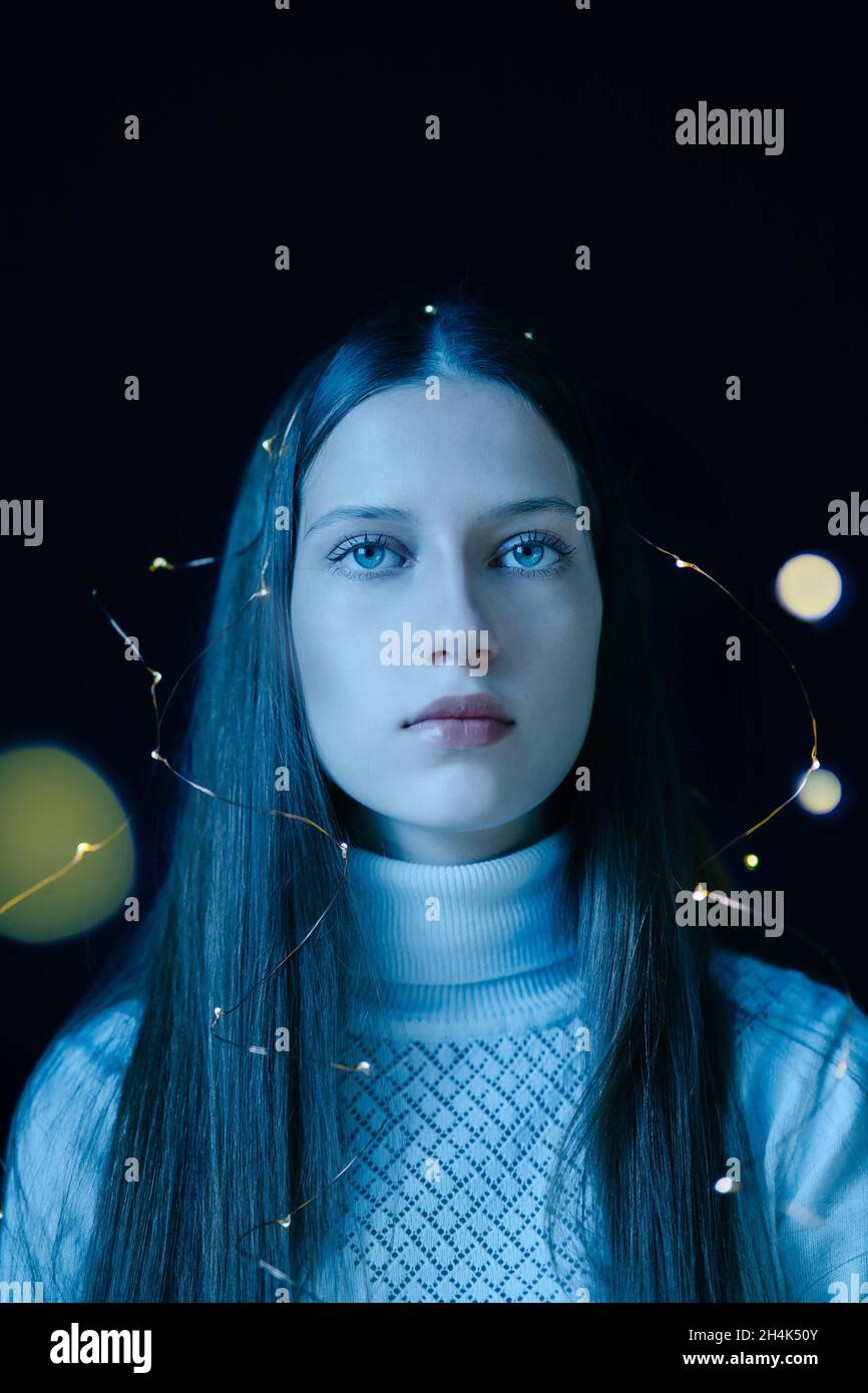Girl with expressive blue eyes on a dark background with garland lights ...
