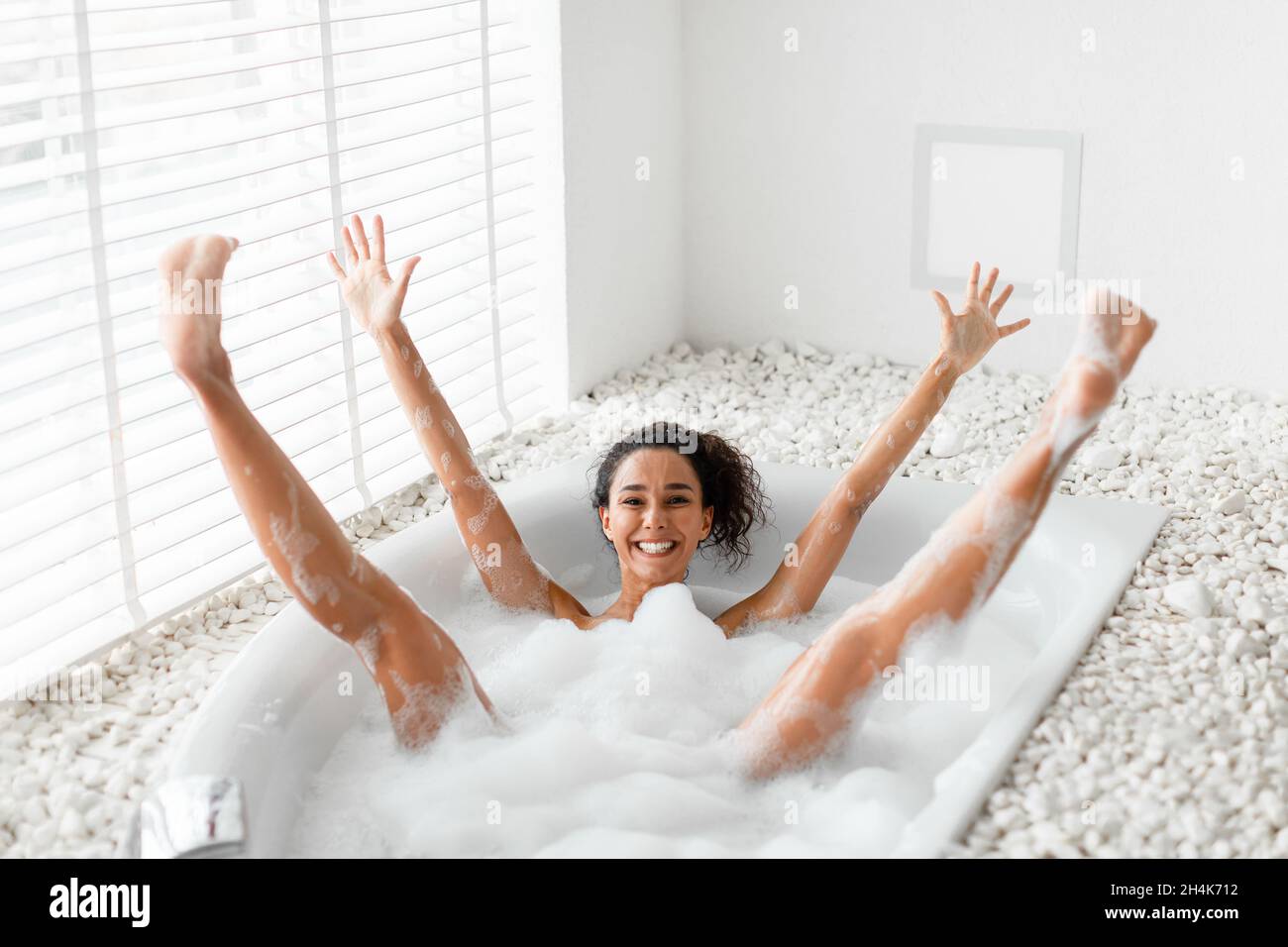 Silly young woman having fun in bubble bath, raising her arms and legs ...
