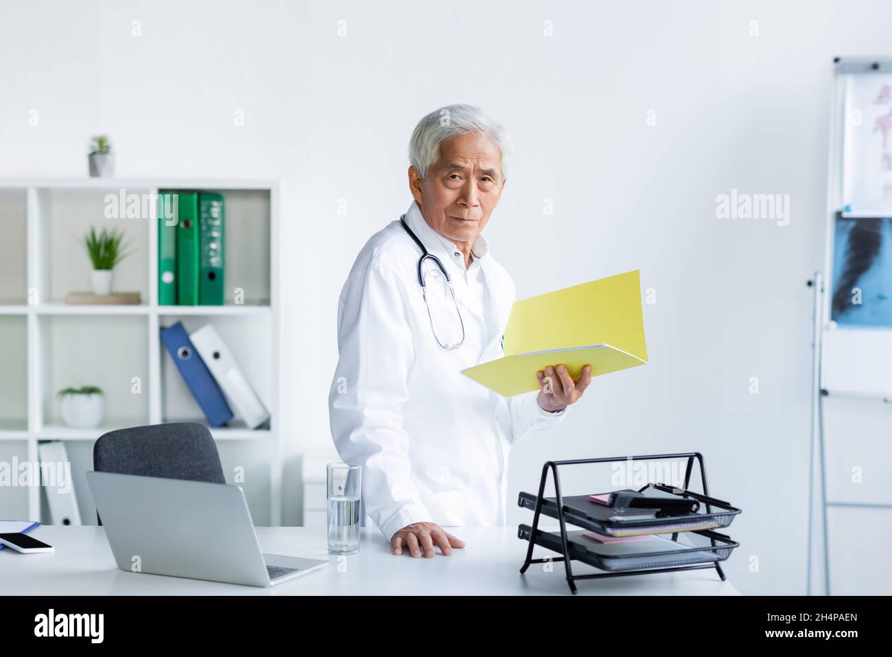 Senior asian doctor holding paper folder near devices and glass of ...