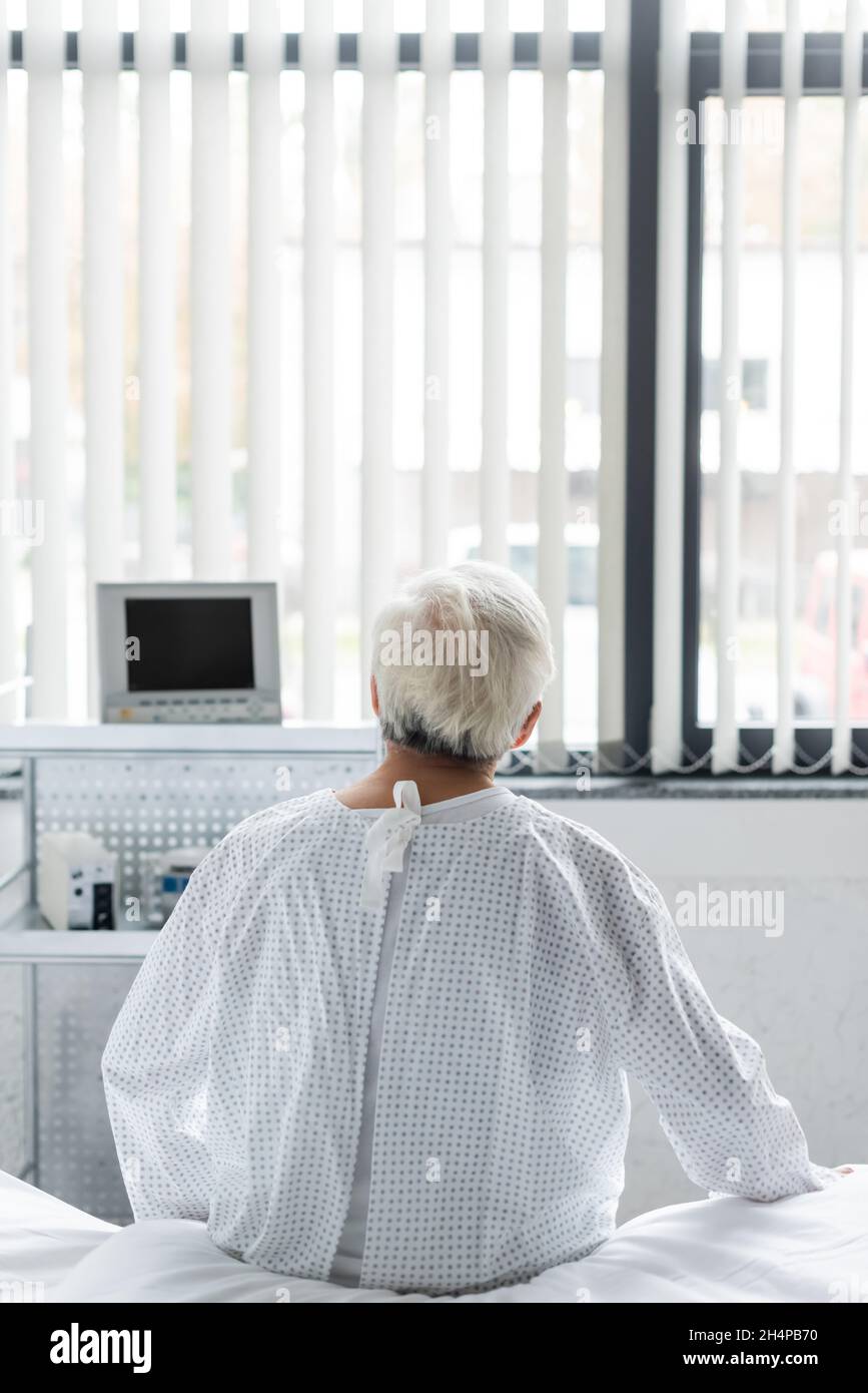 Back view of senior patient in gown sitting on bed in hospital ward ...