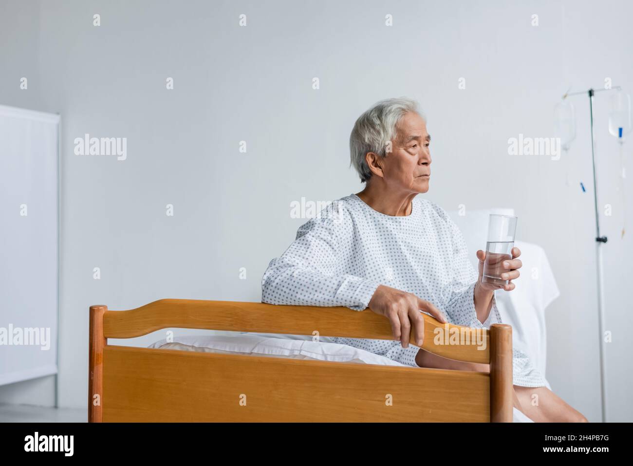Elderly asian patient holding glass of water on bed in hospital ward ...