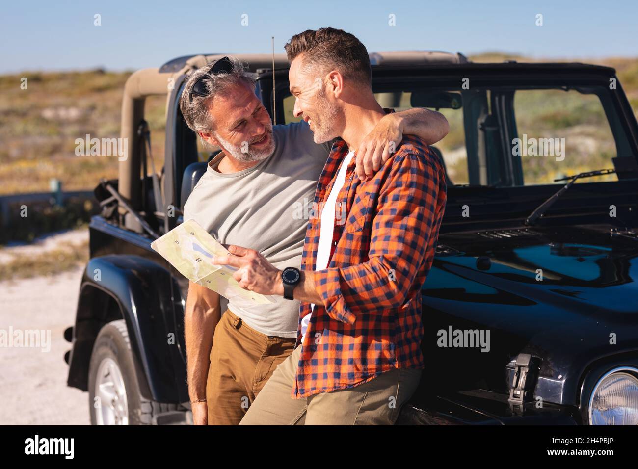 Happy caucasian gay male couple embracing and reading map by car at ...