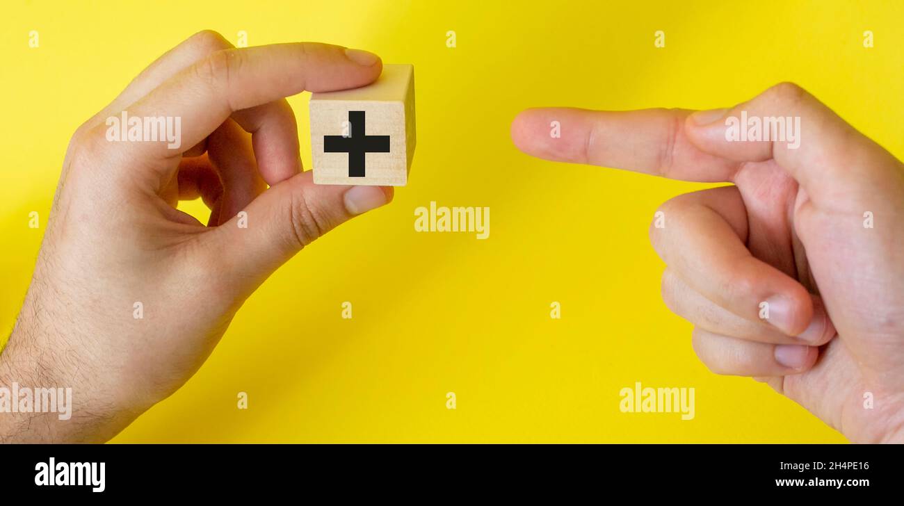 Male hand points to wooden block with PLUS sign on yellow background ...