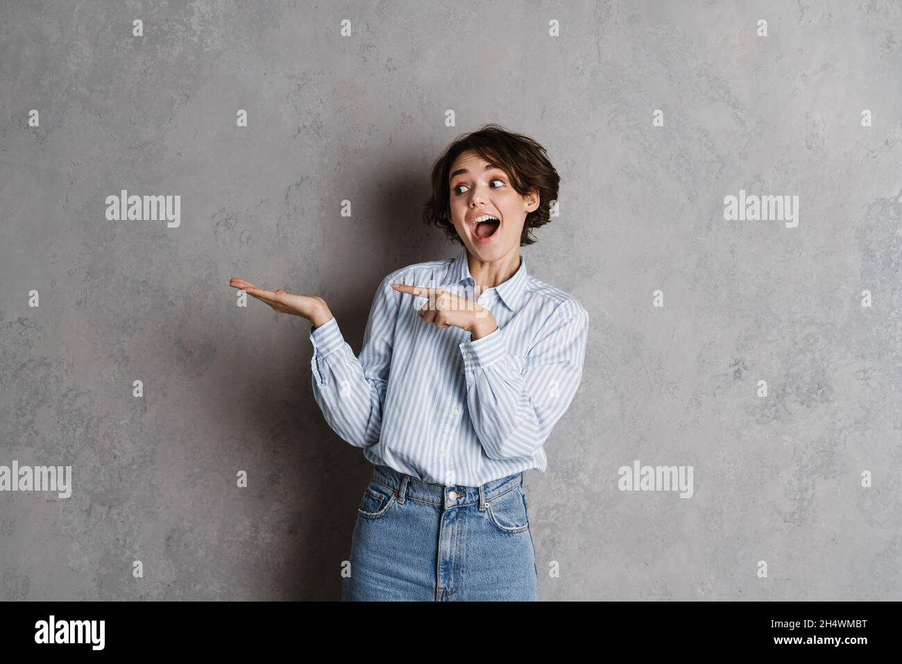 Young brunette woman holding and pointing finger at copyspace isolated over grey wall Stock ...