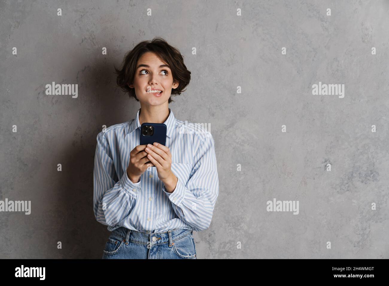 Young brunette woman biting her lip and using mobile phone isolated ...