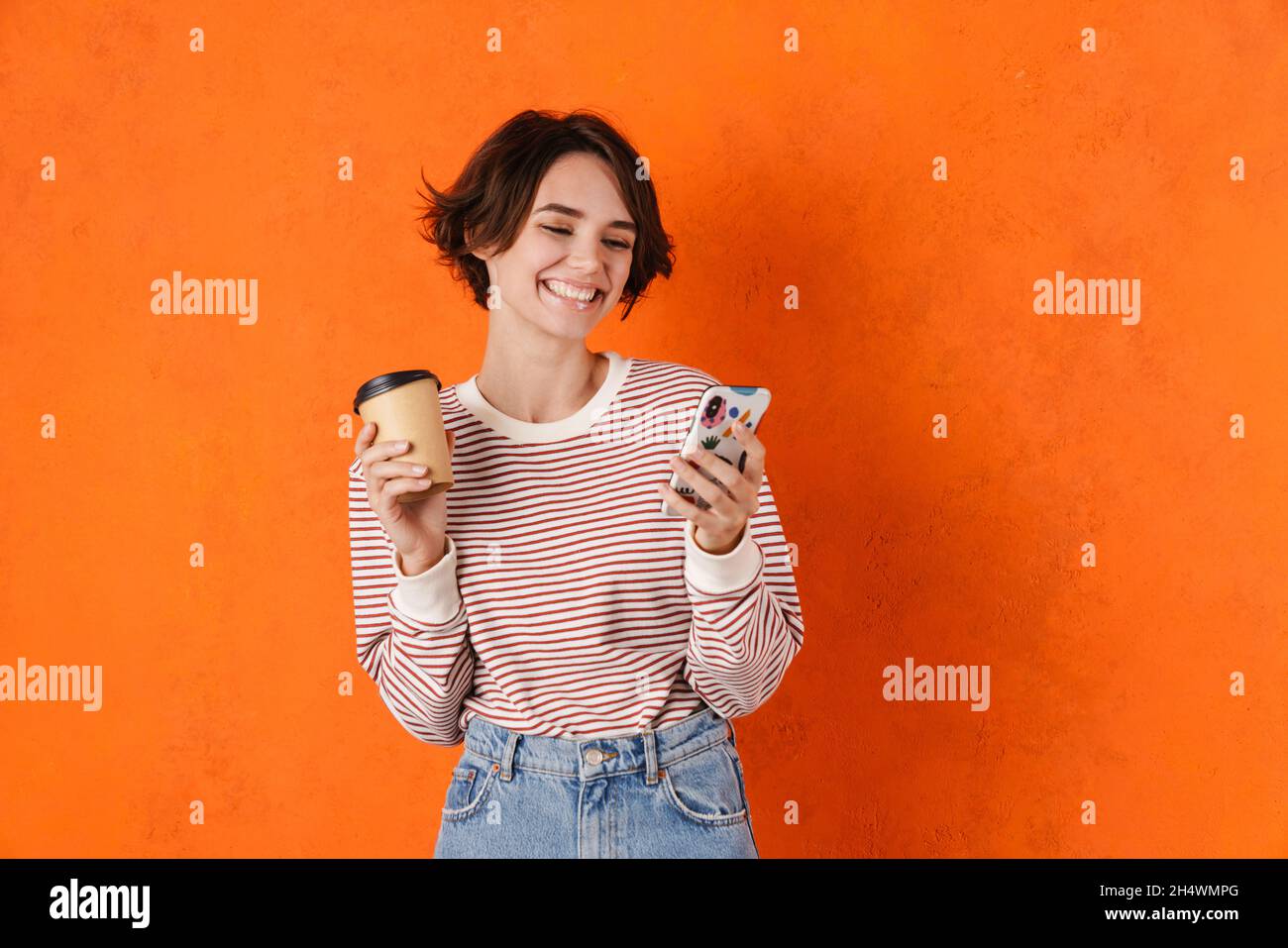 Young brunette woman drinking coffee while using mobile phone isolated over orange wall Stock ...