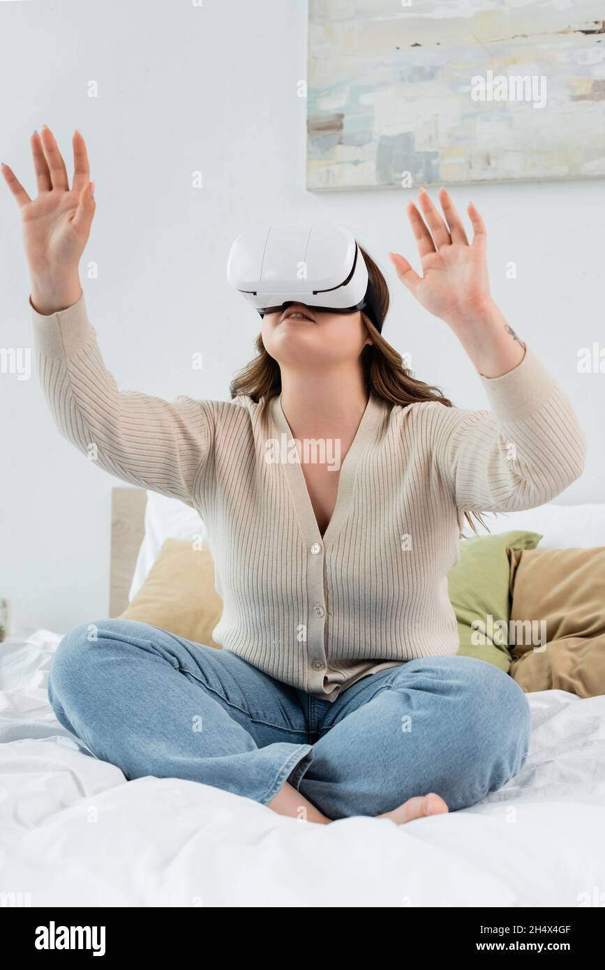 Young body positive woman gaming in vr headset while sitting on bed ...