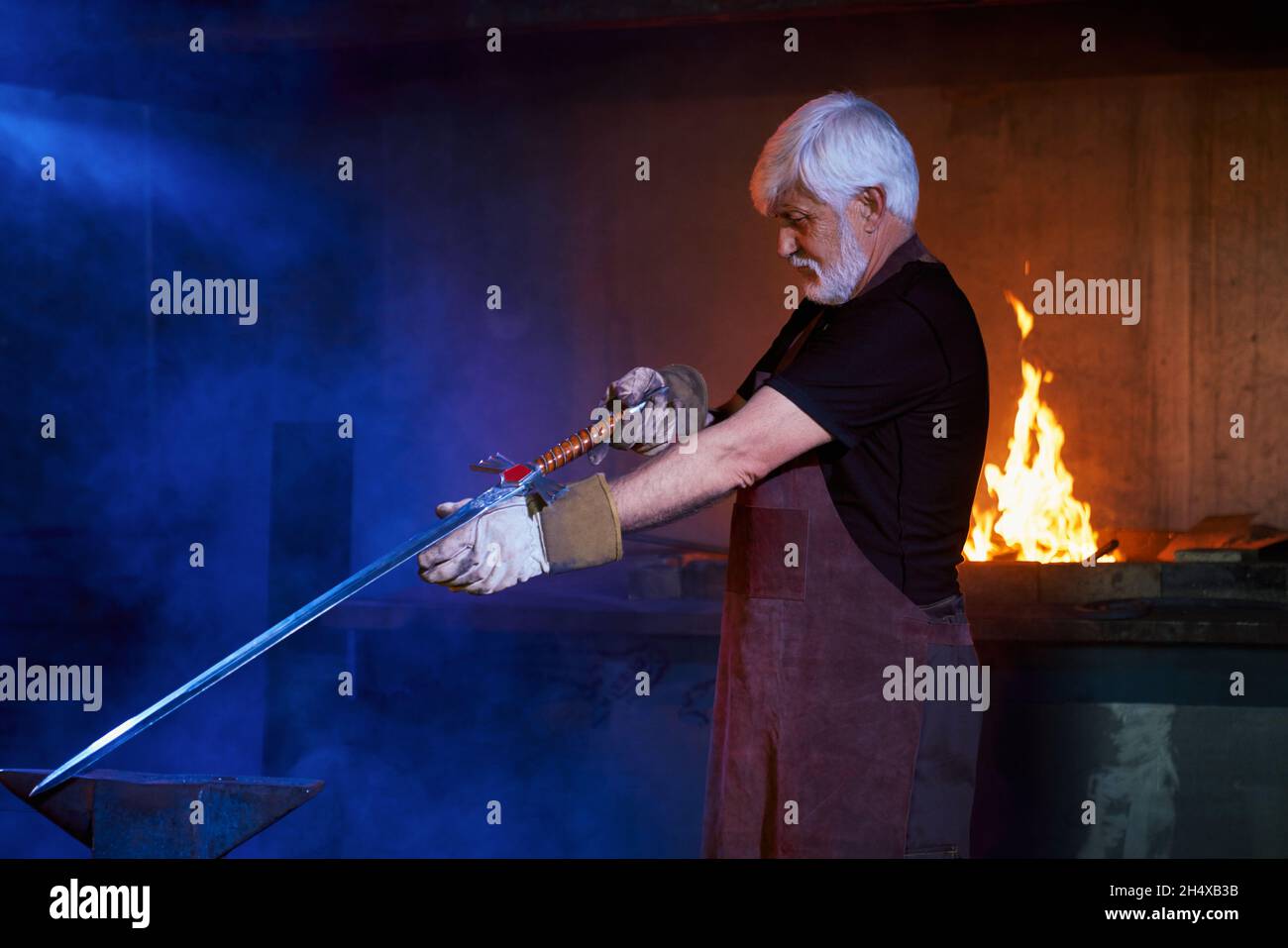 Competent aged blacksmith in safety apron and gloves making sword at ...