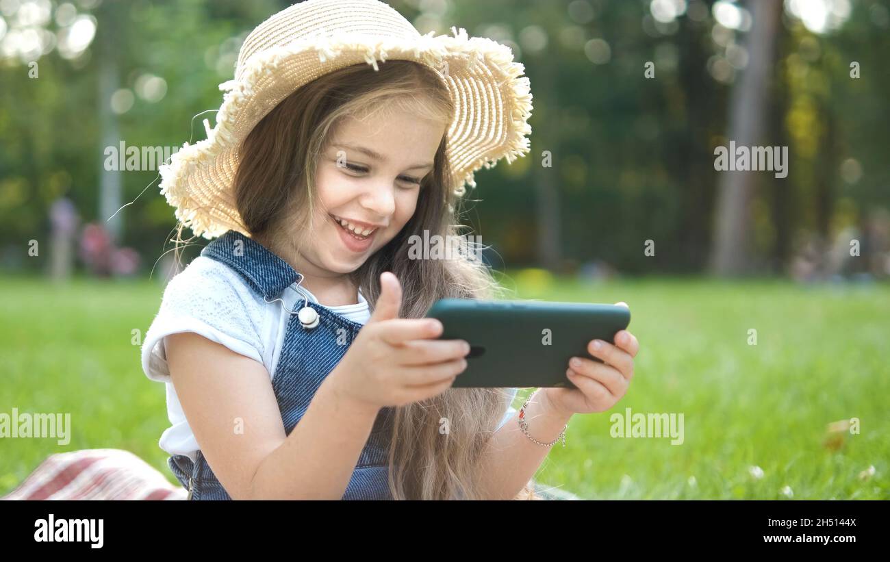 Happy smiling child girl looking in mobile phone outdoors in summer ...