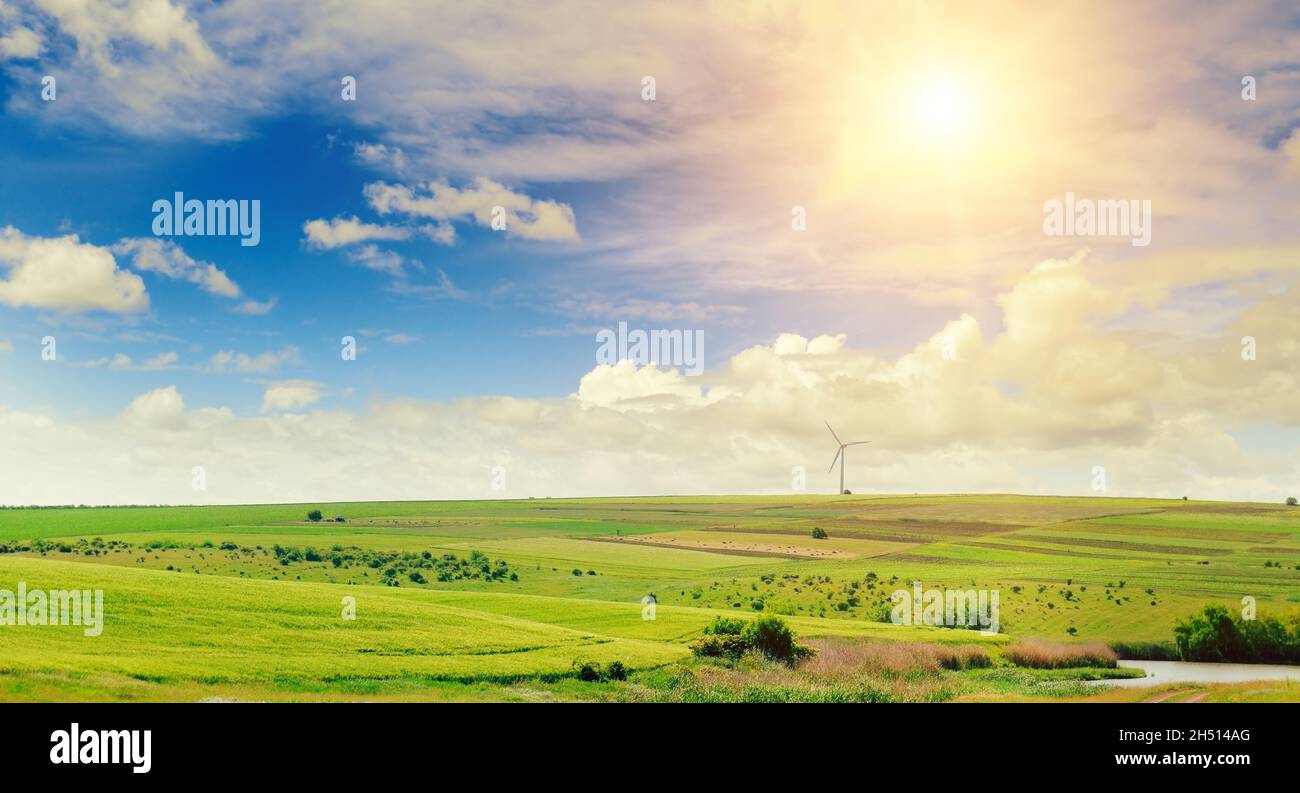 Hilly green field, windmill and sun on blue sky background ...