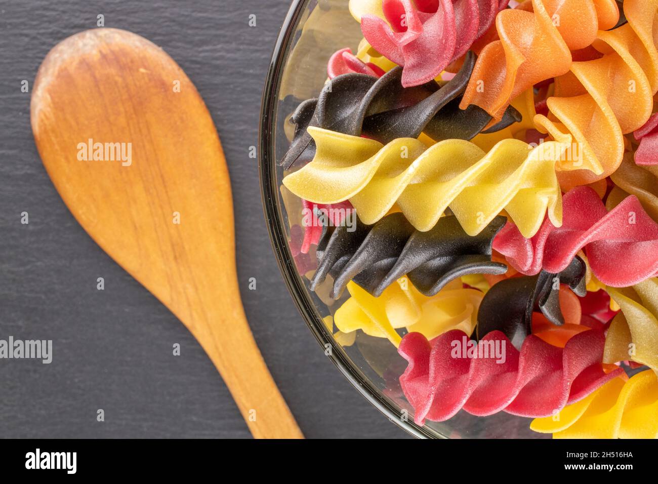 Raw colored fusilli pasta in glass dish with wooden spoon on slate ...