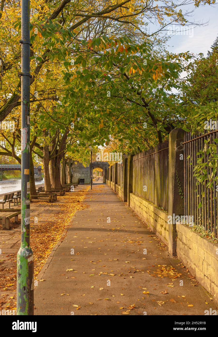 A path runs along a river bank towards an arch. Autumnal trees are ...