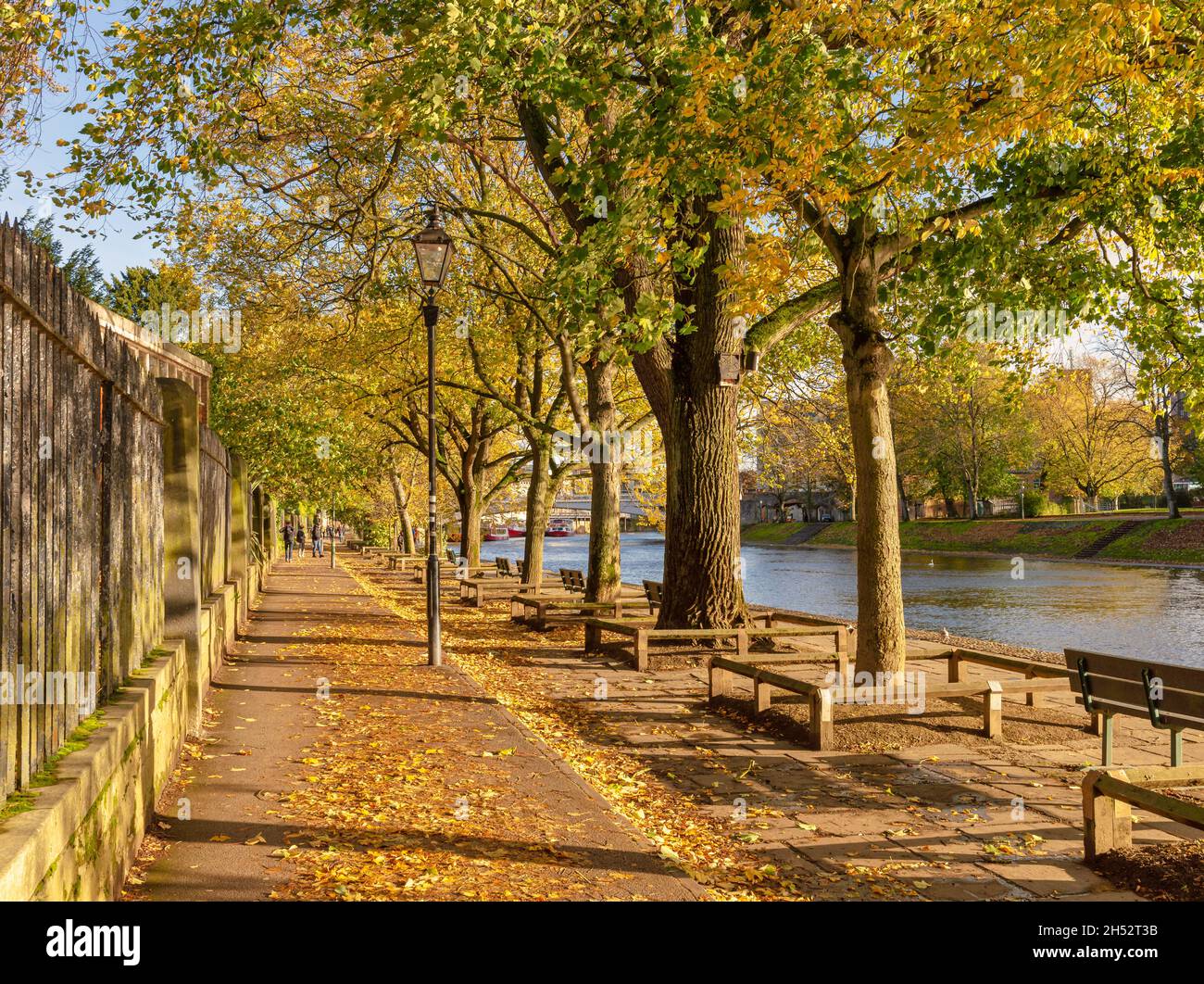 A path runs along a river bank with railings on one side. Autumnal ...