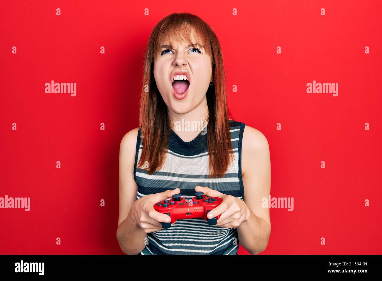 Redhead young woman playing video game holding controller angry and mad ...