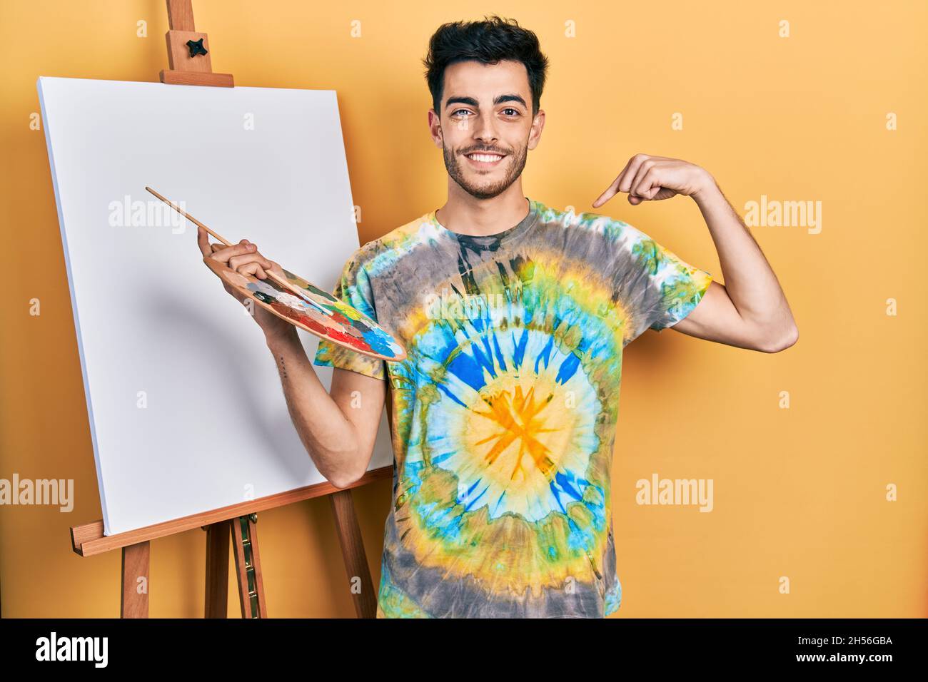 Young hispanic man standing drawing with palette by painter easel stand ...