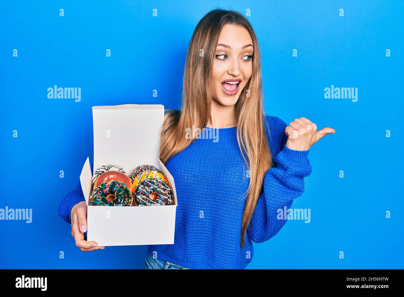 Young blonde girl holding box of tasty colorful doughnuts pointing ...