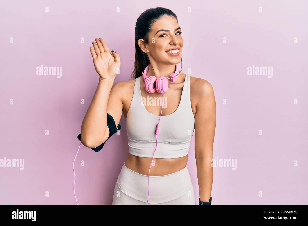 Young hispanic woman wearing gym clothes and using headphones waiving ...