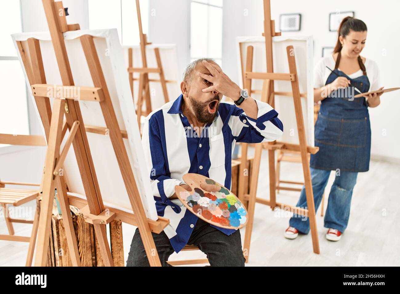Senior artist man at art studio peeking in shock covering face and eyes ...